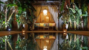 Um jardim interno tranquilo com um lago refletor, uma área de estar com almofadas e uma fonte de luz acolhedora.