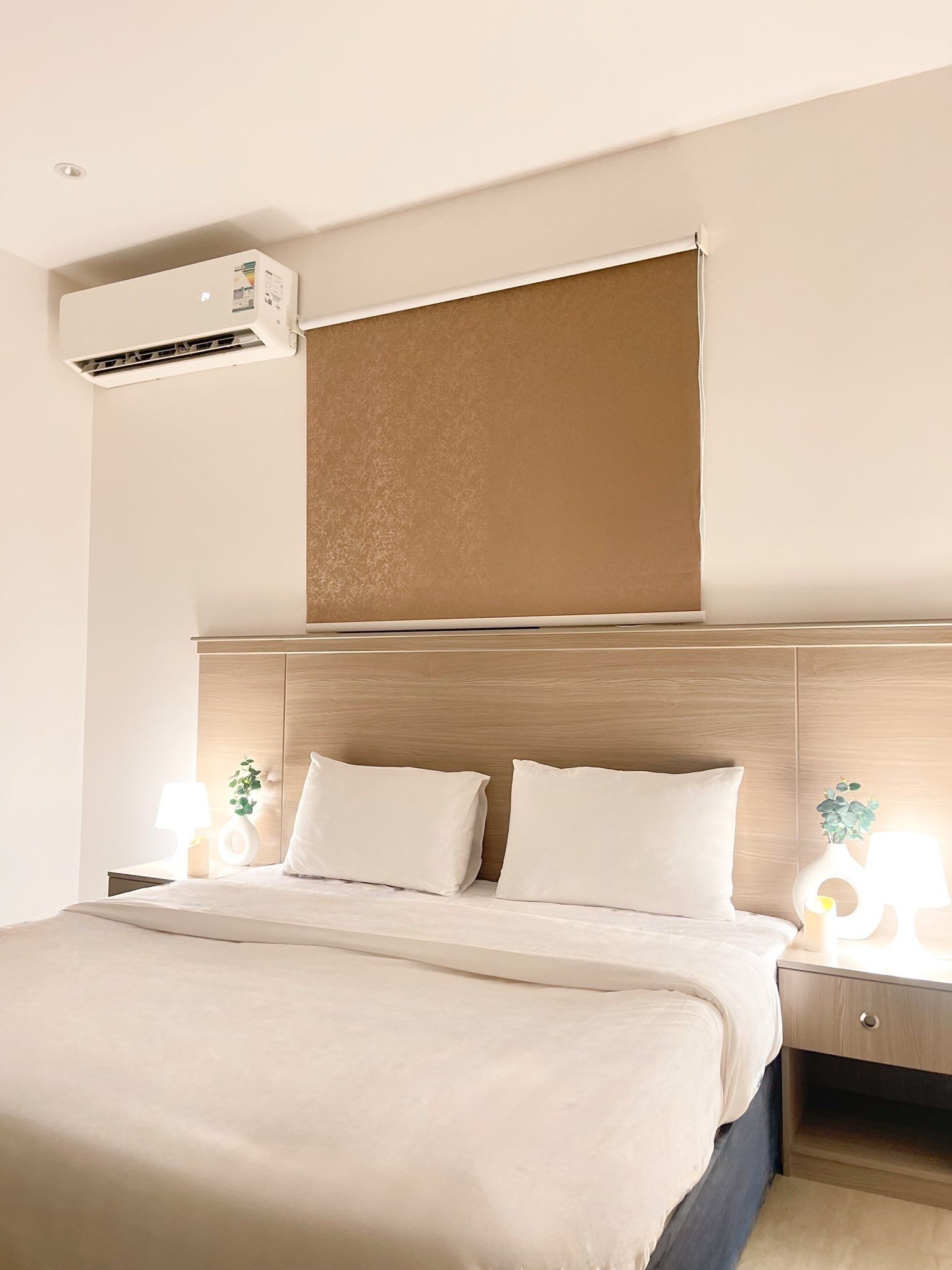 Quarto de hotel com cama, mesas de cabeceira, ar condicionado fixado na parede e uma janela grande com persiana marrom.