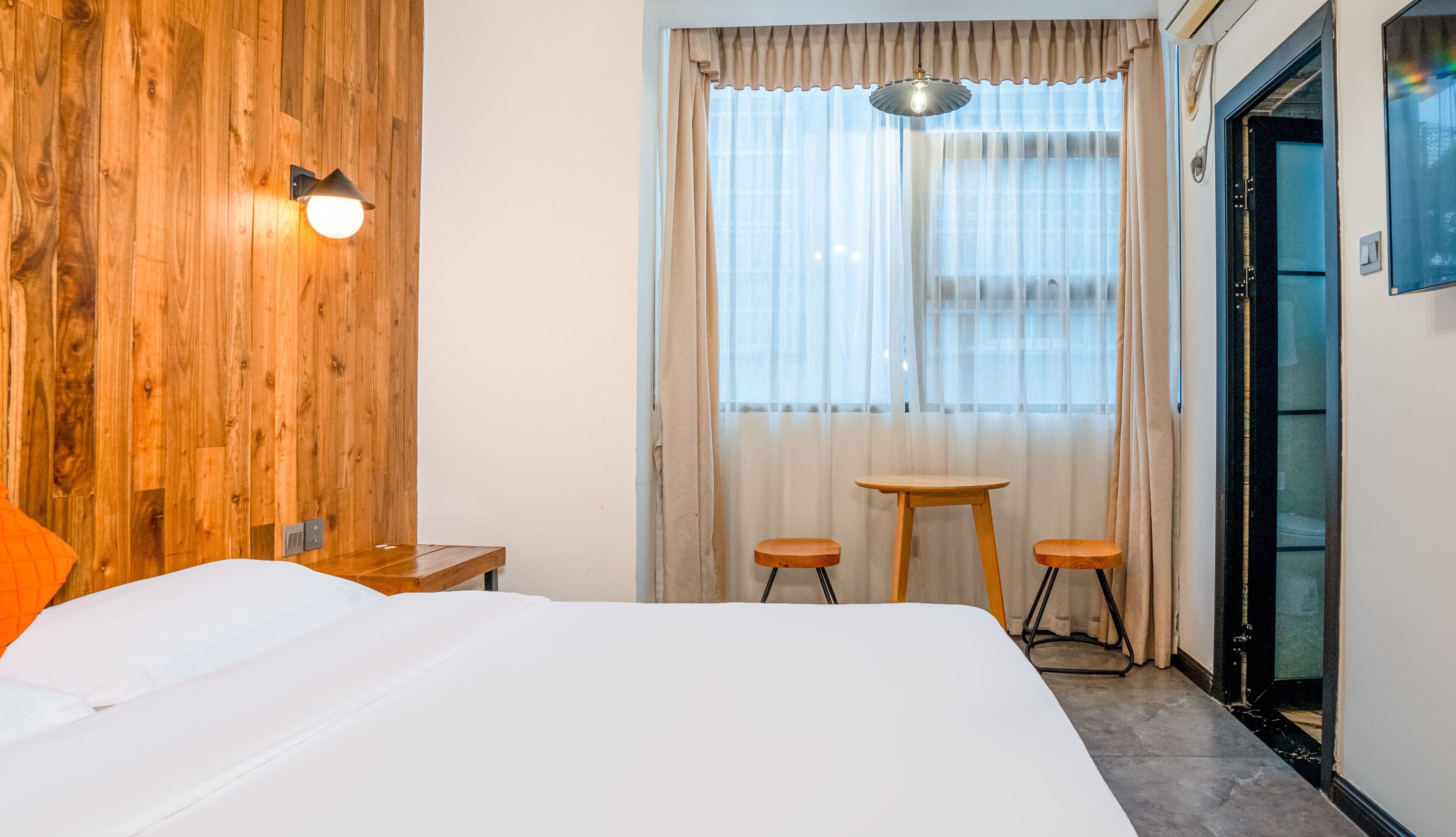 Quarto de hotel com parede de madeira, uma cama, uma escrivaninha, dois banquinhos, uma janela com cortinas e uma porta.