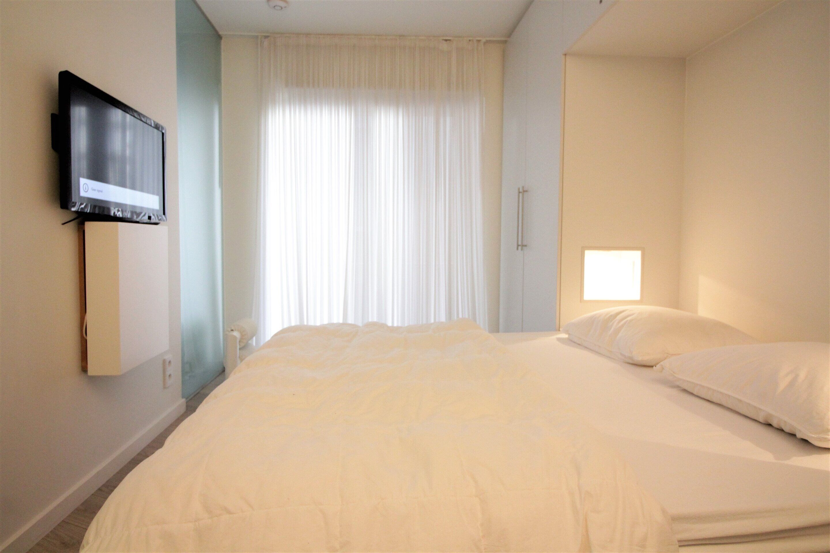 Quarto de hotel com cama, televisão na parede, janela com cortinas e pequena mesa de cabeceira.