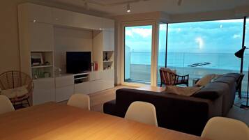 Uma sala de estar moderna com uma mesa de jantar de madeira, cadeiras brancas, um sofá e uma televisão sobre um móvel branco. Há uma janela grande que oferece vista para o mar.