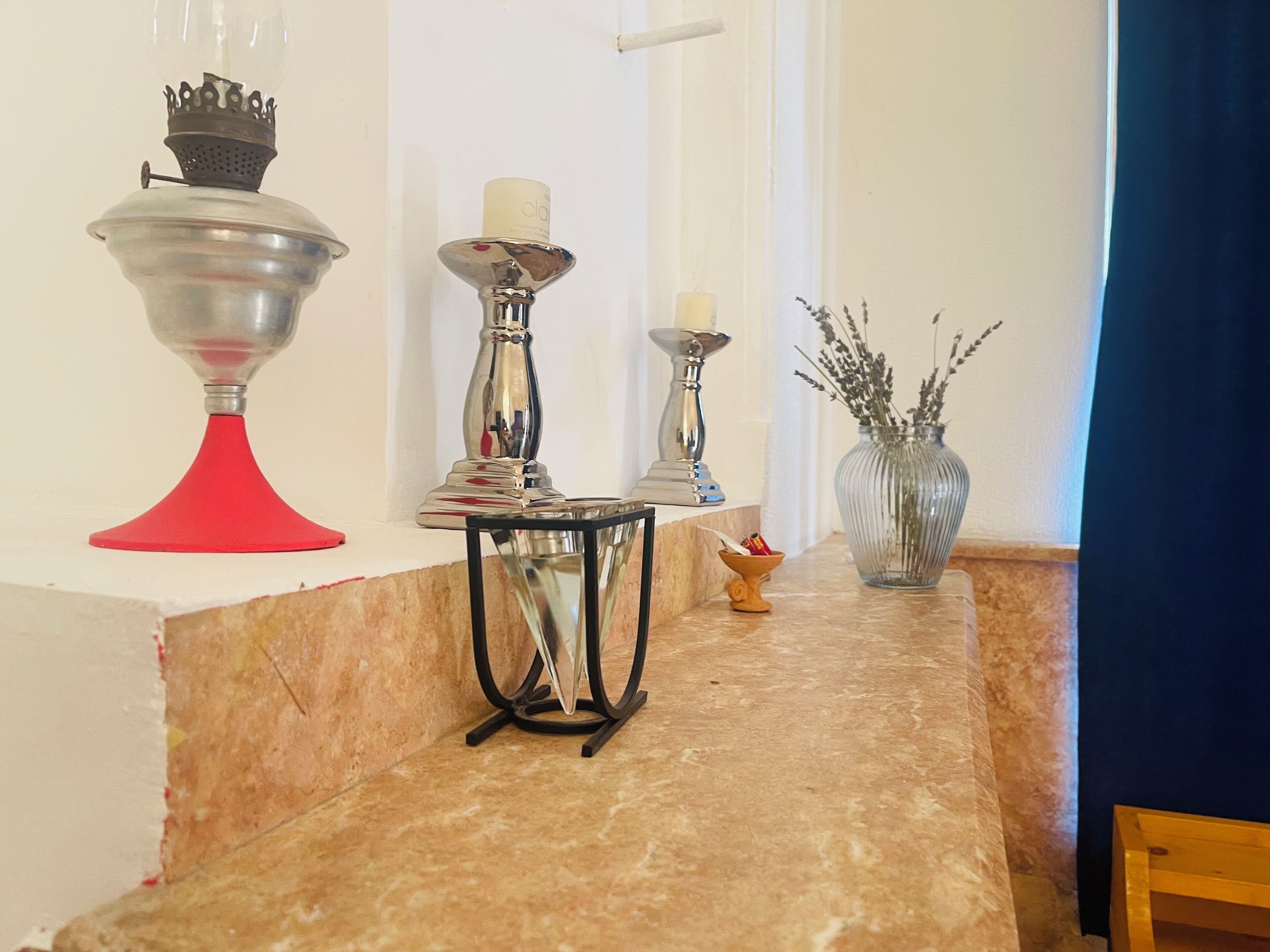 Um corredor revestido de mármore, com um candeeiro pedestal vermelho, uma pequena mesa com uma vela e um vaso com flores secas.