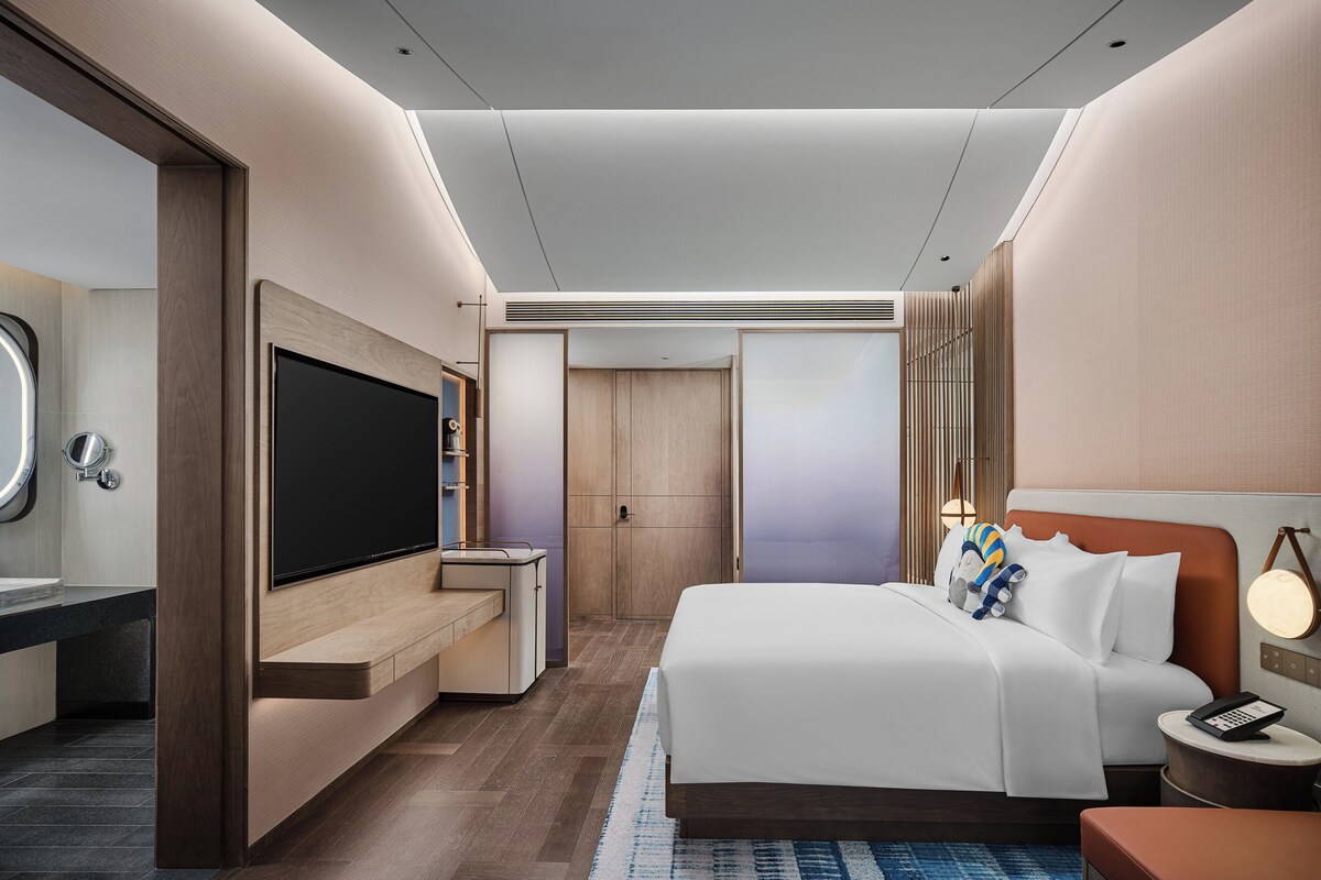 Quarto de hotel moderno com cama grande, mesa de cabeceira, televisão e banheiro com espelho e pia.