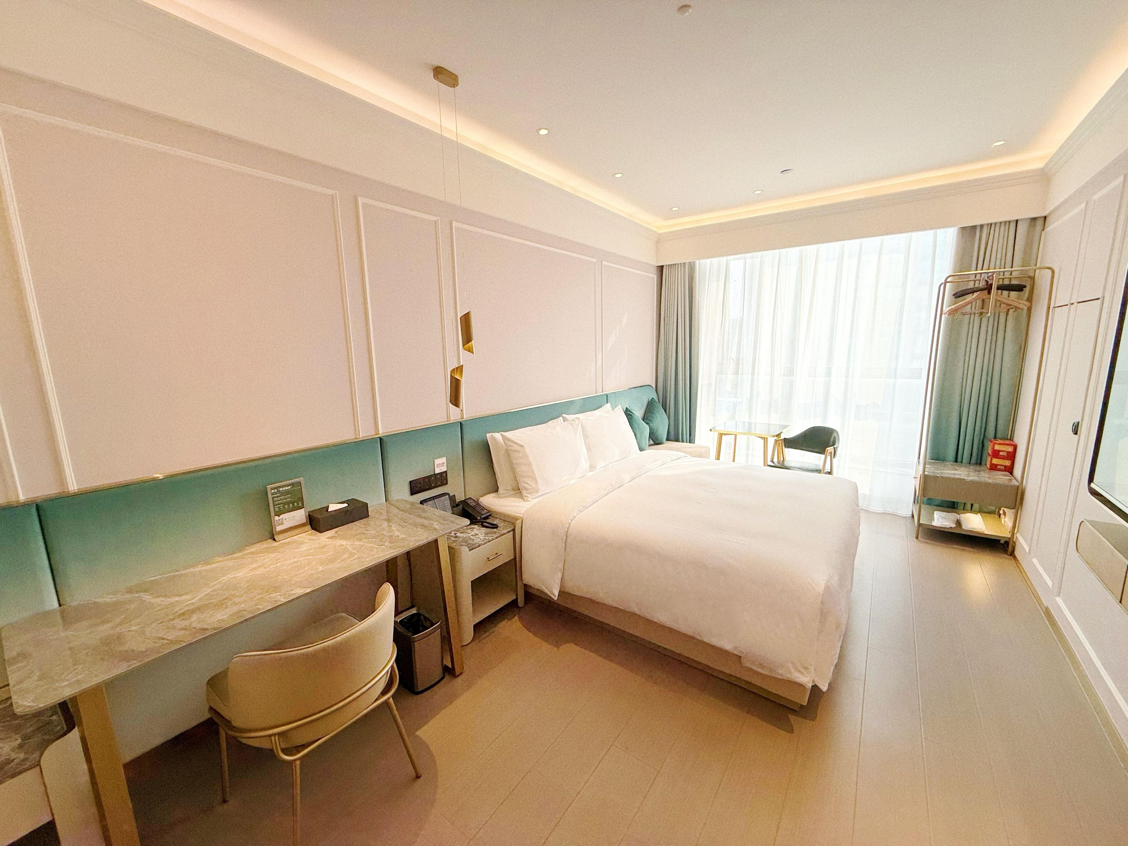Quarto de hotel com cama, escrivaninha, cadeira, mesa pequena, espelho e guarda-roupa.