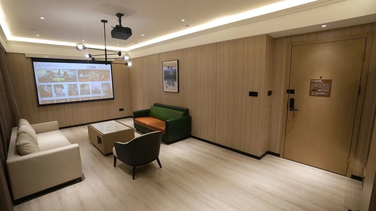 Quarto de hotel moderno com uma tela grande, um sofá, uma cadeira, uma mesinha e uma porta de madeira.
