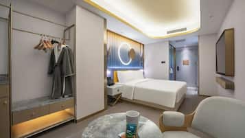 Quarto de hotel moderno com cama, mesa de cabeceira, mesa de centro redonda, área de estar, guarda-roupa com roupas penduradas e luminária circular na parede.