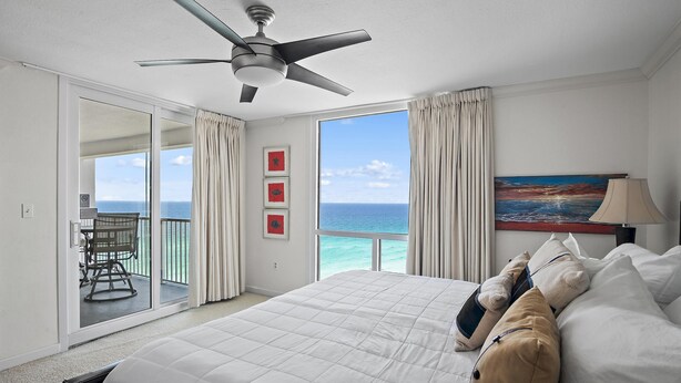 Quarto de hotel com cama, ventilador de teto, varanda com mesa e cadeiras, e um quadro na parede.