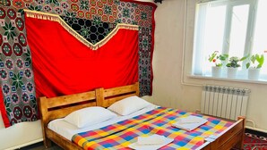 Uma cama de madeira com colcha xadrez colorida, almofadas brancas e cortina vermelha com padrão geométrico.
