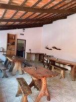 Uma área de refeições ao ar livre rústica, com mesas e bancos de madeira, uma lareira de tijolos e piso de azulejos.