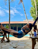 Uma pessoa relaxando em uma rede perto de uma piscina com um pátio ladrilhado e palmeiras.