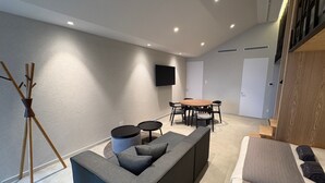 Uma sala de estar moderna com sofá, uma mesa de jantar com cadeiras, uma TV fixada na parede e uma escada que leva a um andar superior.
