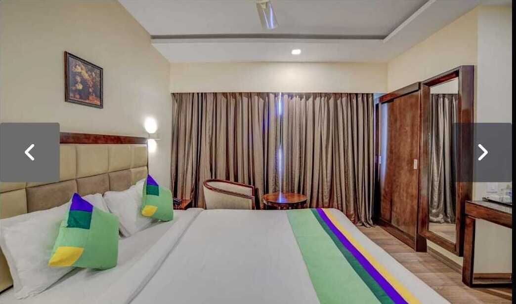 Quarto de hotel com cama, cadeira, escrivaninha e uma janela grande com cortinas.