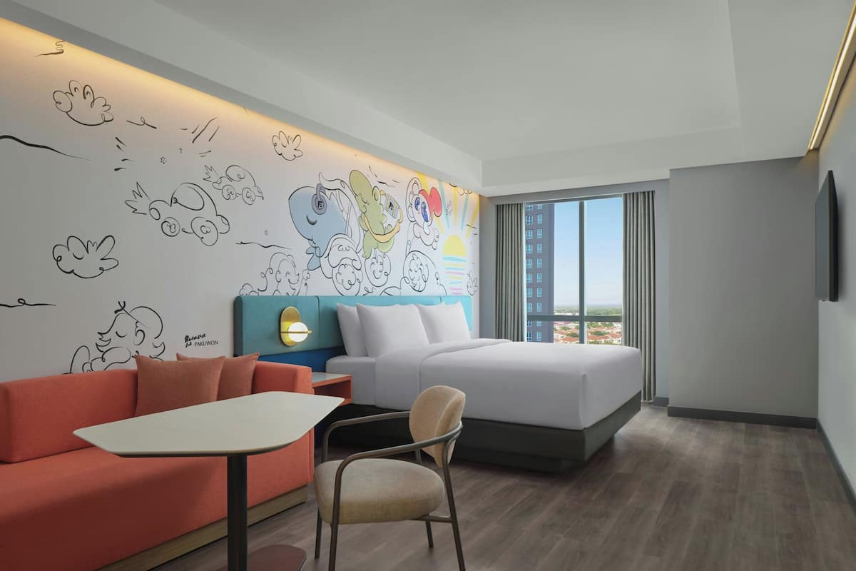 Quarto de hotel moderno com uma cama grande, um sofá, uma mesa pequena e uma cadeira. Há um mural na parede com personagens de desenho animado e uma grande janela com vista para a cidade.