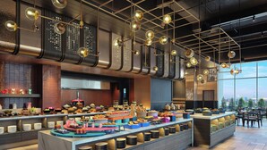 Disposição moderna de buffet com diversos tipos de comida, teto alto com luminárias pendentes e vista para a cidade através de grandes janelas.