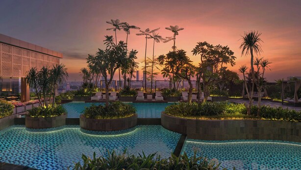 Piscina no terraço com palmeiras, espreguiçadeiras e vista para a cidade ao pôr do sol.