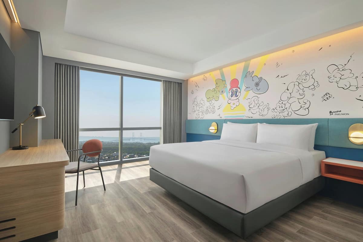 Quarto de hotel moderno com uma cama grande, uma escrivaninha, uma cadeira e vista da cidade através de janelas do chão ao teto.