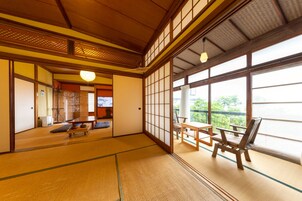 Um quarto japonês tradicional com tatames, uma porta deslizante e vista para o verde lá fora, e uma mesa com cadeiras.