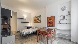 Quarto compacto com cama, mesa de jantar com cadeiras, relógio fixado na parede e prateleiras com objetos decorativos.