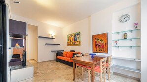 Uma sala de estar moderna com sofá, mesa de jantar e cadeiras. Há uma escada com prateleira, um relógio de parede e obras de arte.