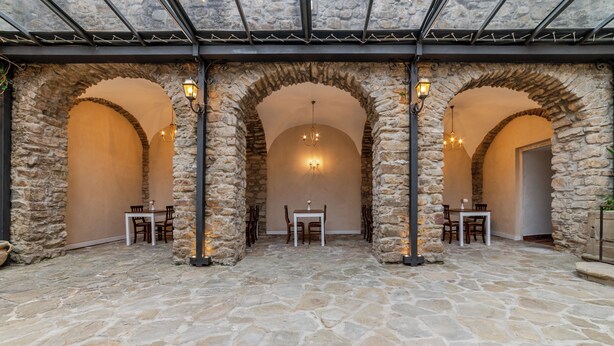 Pátio de pedra com arcos, mesas e cadeiras, e um lustre pendurado.