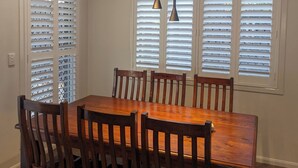 Uma sala de jantar com mesa e cadeiras de madeira, três luminárias pendentes e janelas com moldura branca e persianas.
