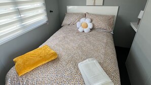Uma cama bem arrumada com um cobre-leito estampado, um travesseiro grande com motivos florais e uma toalha amarela.