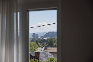 Visão através de uma janela com céu claro, edifícios e uma colina distante.