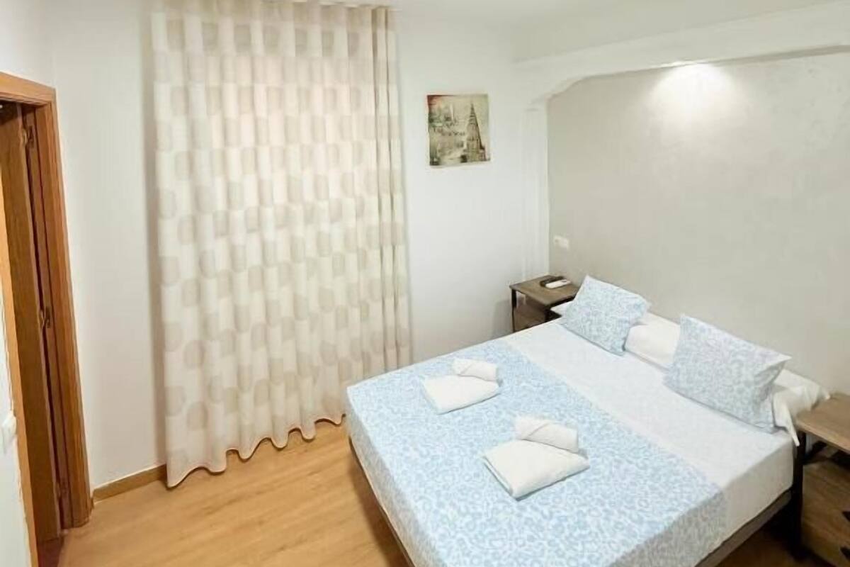 Quarto com cama, mesa de cabeceira, luminária na parede, janela com cortinas e porta.