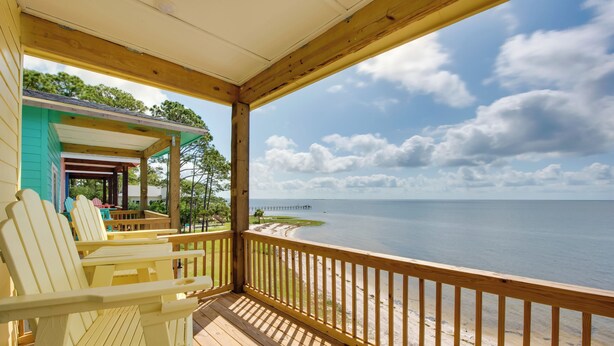 Um deck de madeira com cadeiras Adirondack com vista para uma praia e oceano.