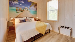 Quarto com teto de madeira, cama com colcha branca e manta amarela, mesa de cabeceira, luminária, janela com persianas e um mural de uma cena de praia.
