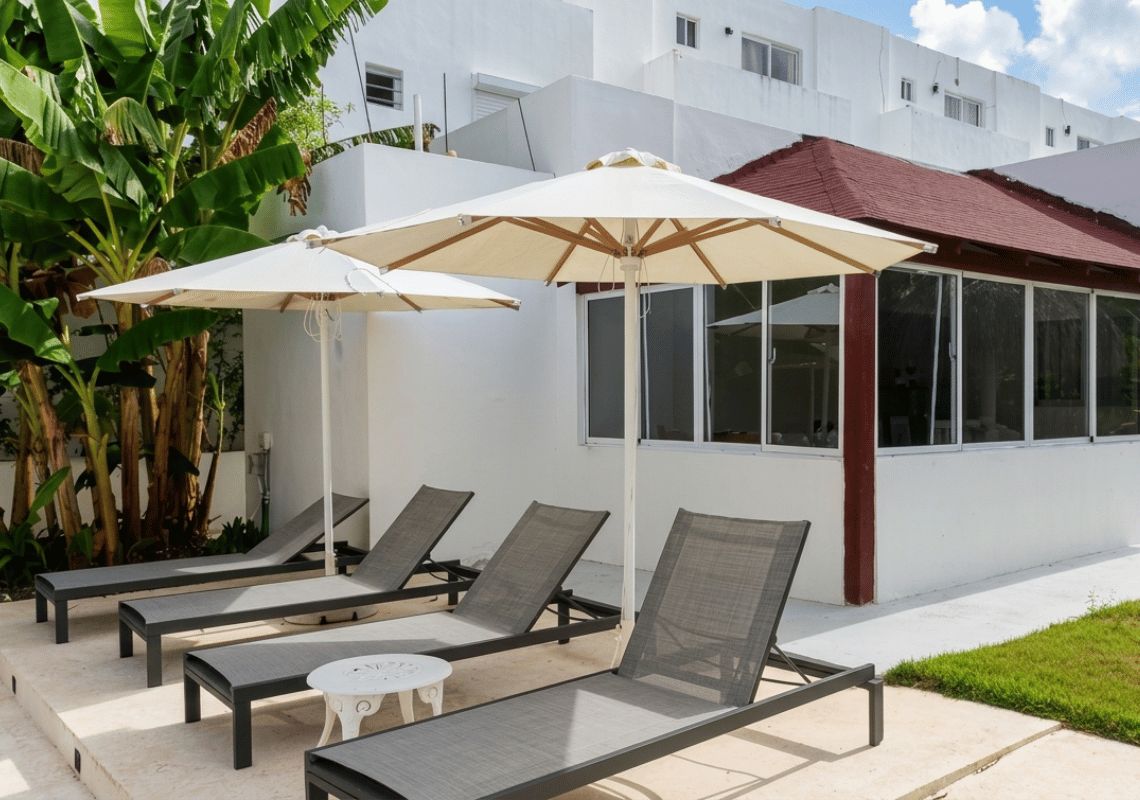 Área da piscina com espreguiçadeiras e guarda-sóis, um edifício branco com janelas amplas e um telhado vermelho.