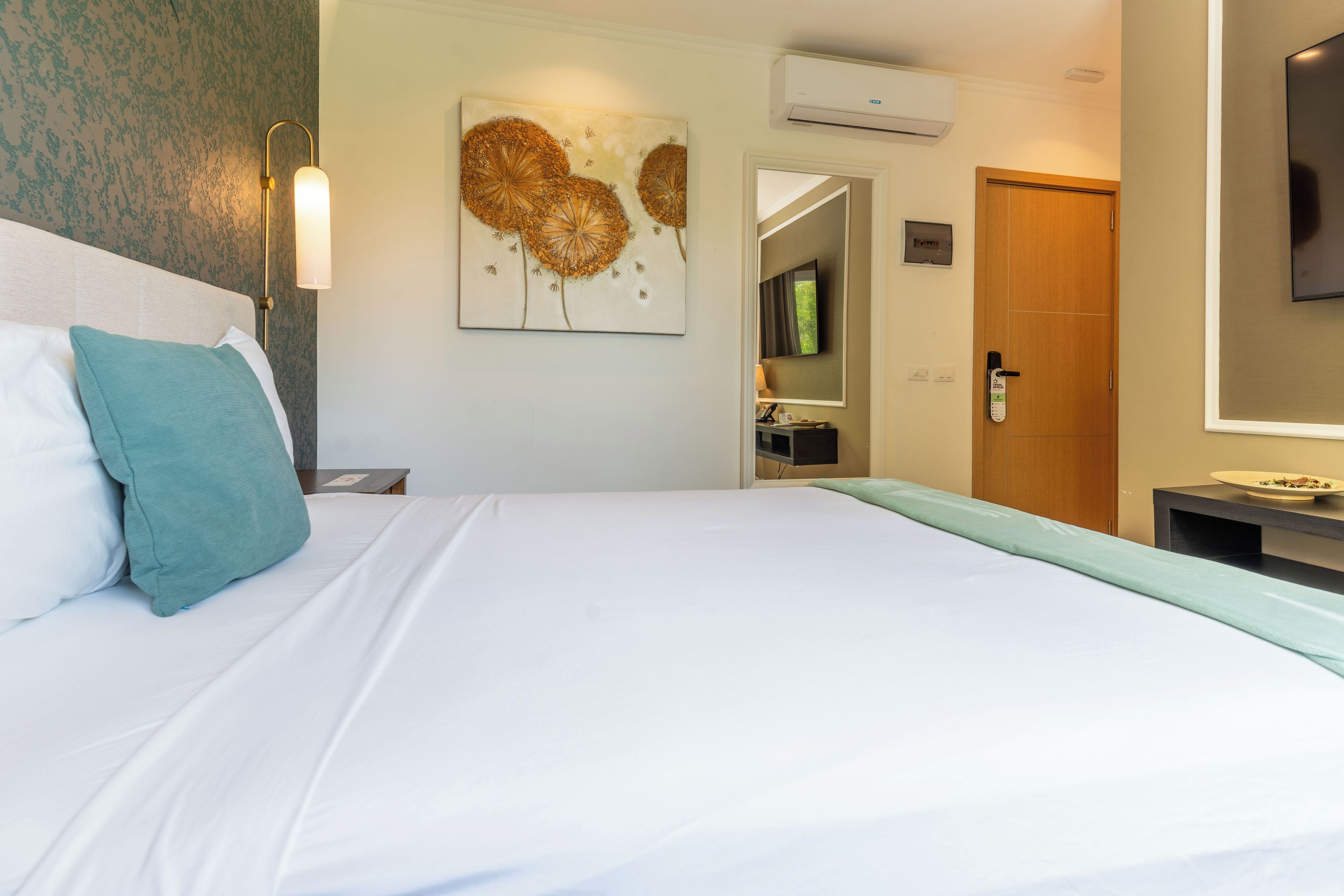 Quarto de hotel com cama, mesa de cabeceira, televisão na parede e porta.