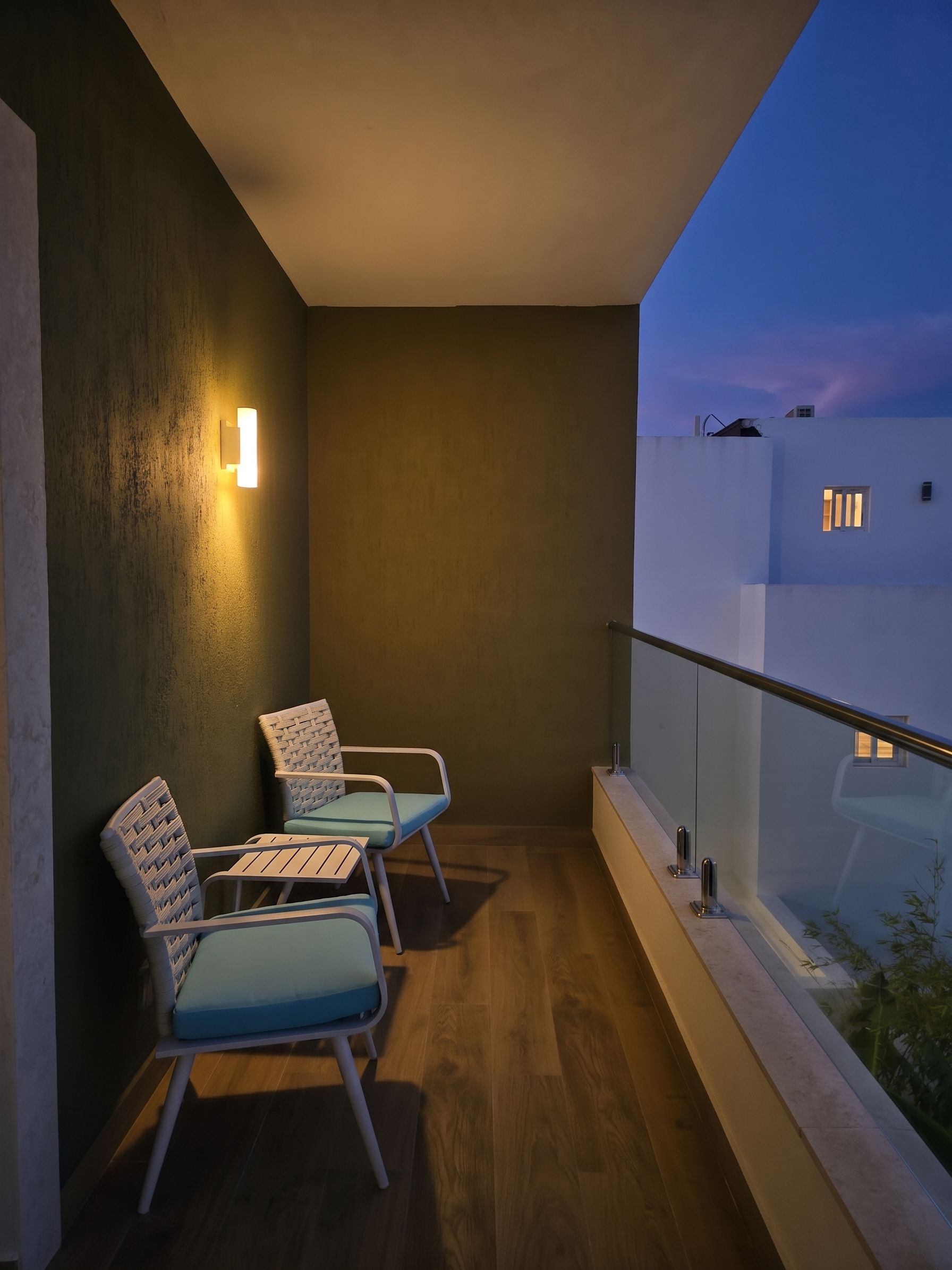 Uma varanda com duas cadeiras e uma mesa, corrimão e vista de edifícios ao fundo.