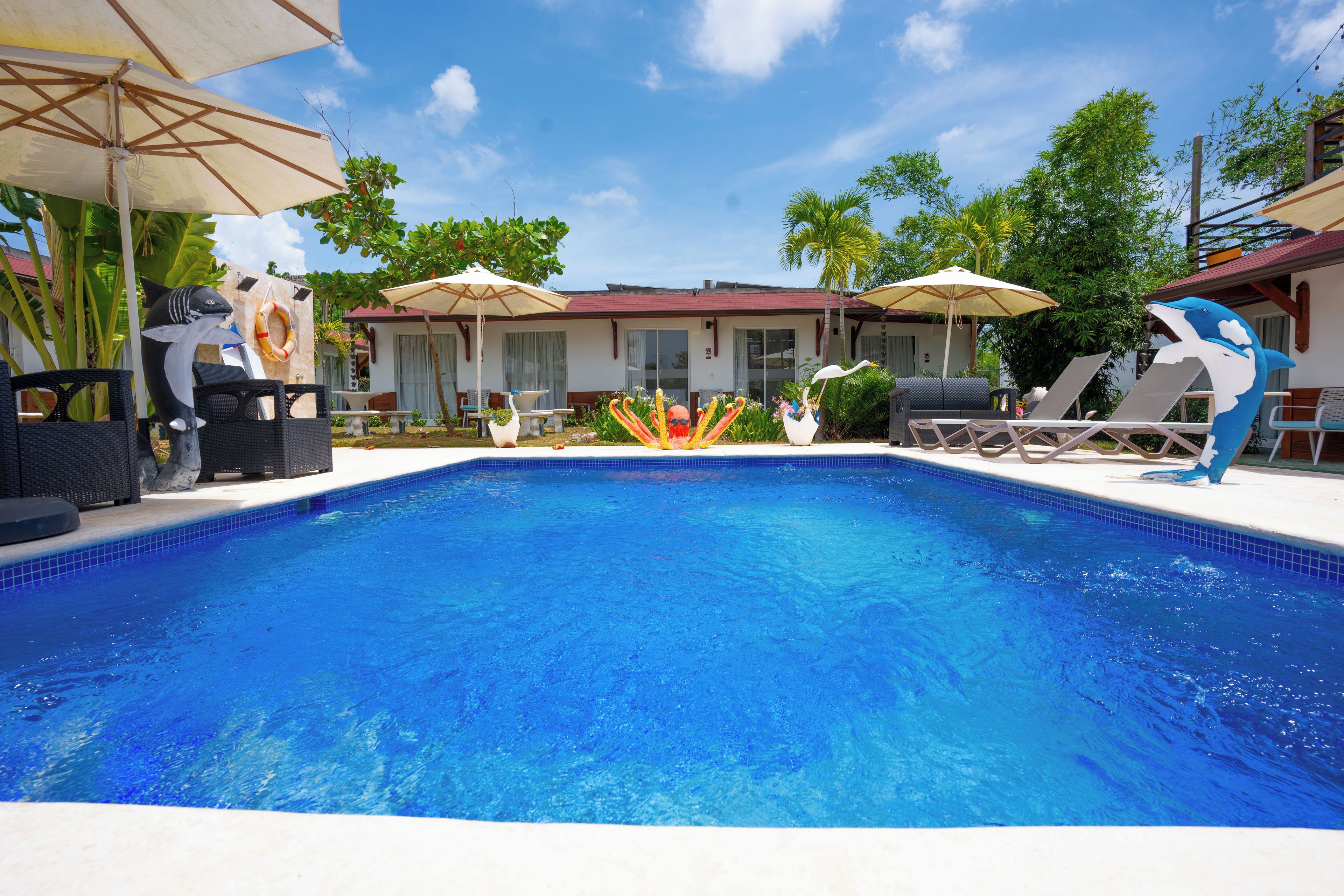 Piscina com espreguiçadeiras e guarda-sóis, cercada por plantas tropicais e um edifício com telhado vermelho.