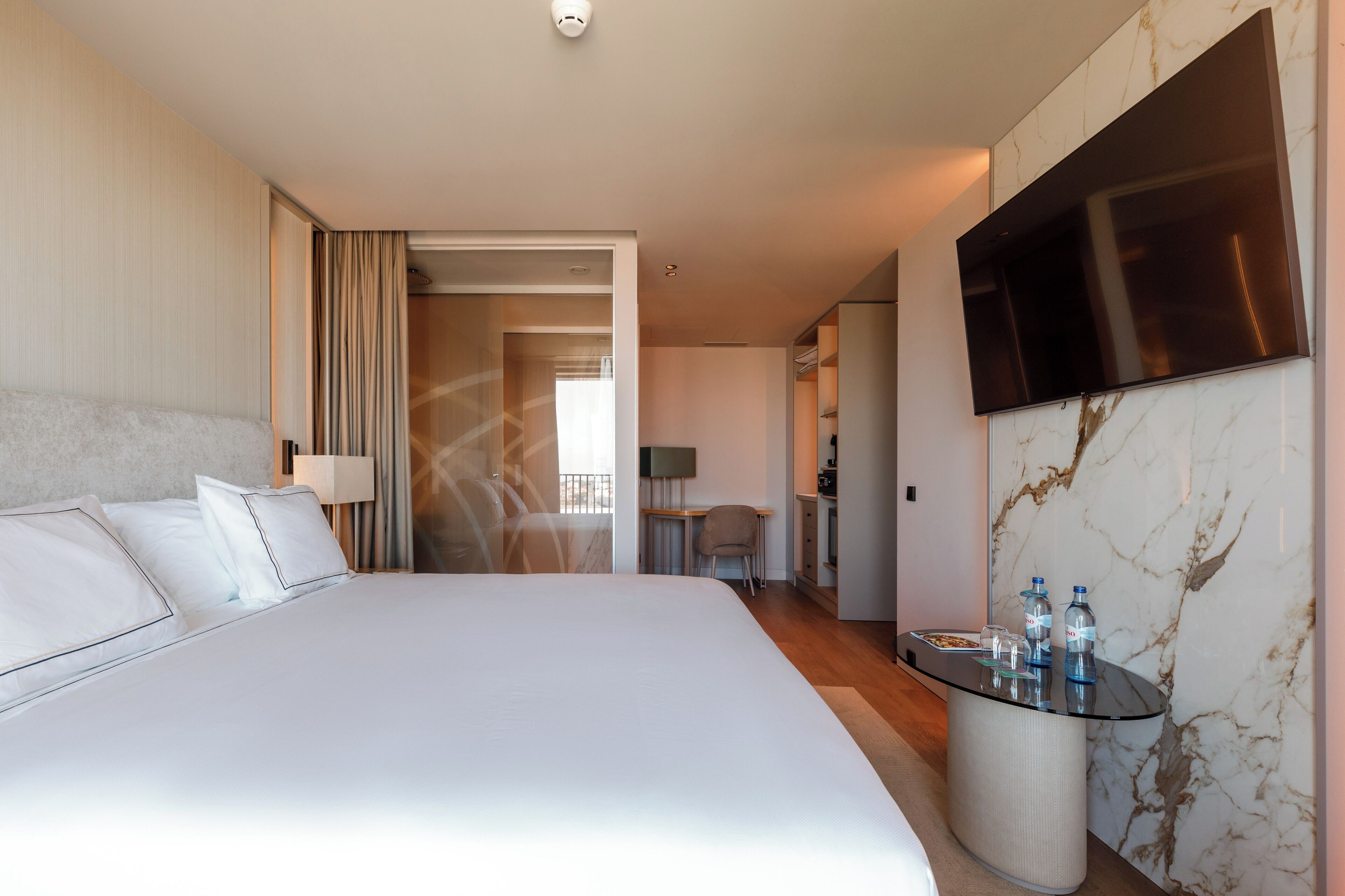 Quarto de hotel com uma cama grande, televisão fixada na parede, escrivaninha com cadeira e uma mesinha com garrafas.