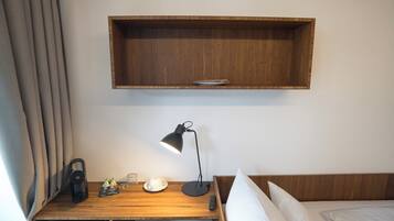 Quarto de hotel com mesa de cabeceira de madeira, luminária de mesa, controle remoto e uma pequena mesa com uma xícara e uma garrafa.