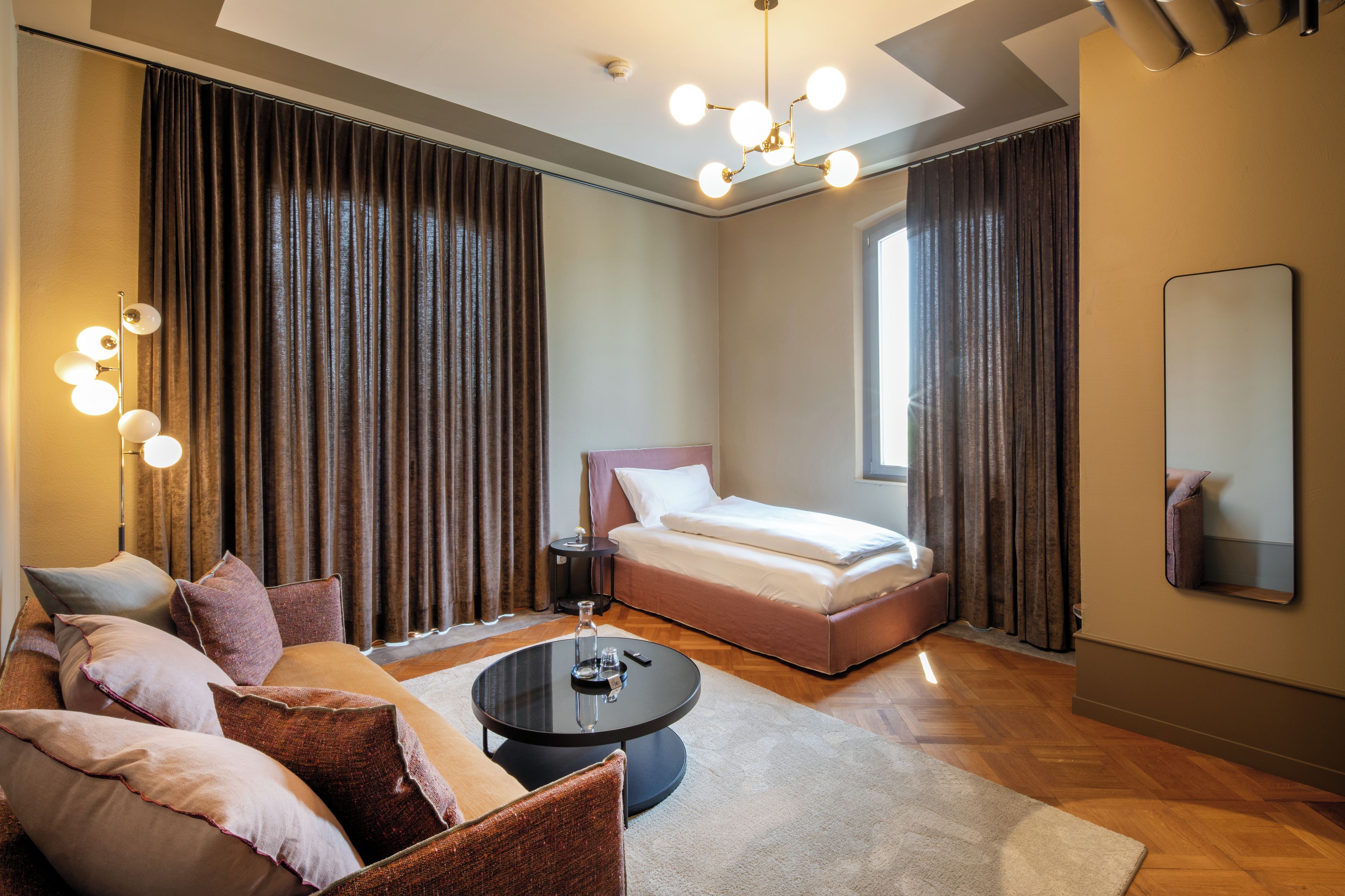Quarto de hotel moderno com cama, sofá, mesa de centro e um espelho grande.
