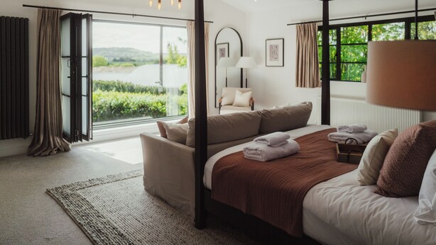 Uma cama com dossel, um edredom marrom, uma janela grande com cortinas e vista para o verde, e uma mesa de cabeceira com uma luminária.