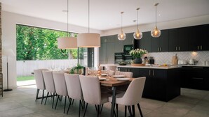 Cozinha moderna com armários escuros, ilha central, iluminação pendente e área de refeições com cadeiras e uma mesa posta para seis.