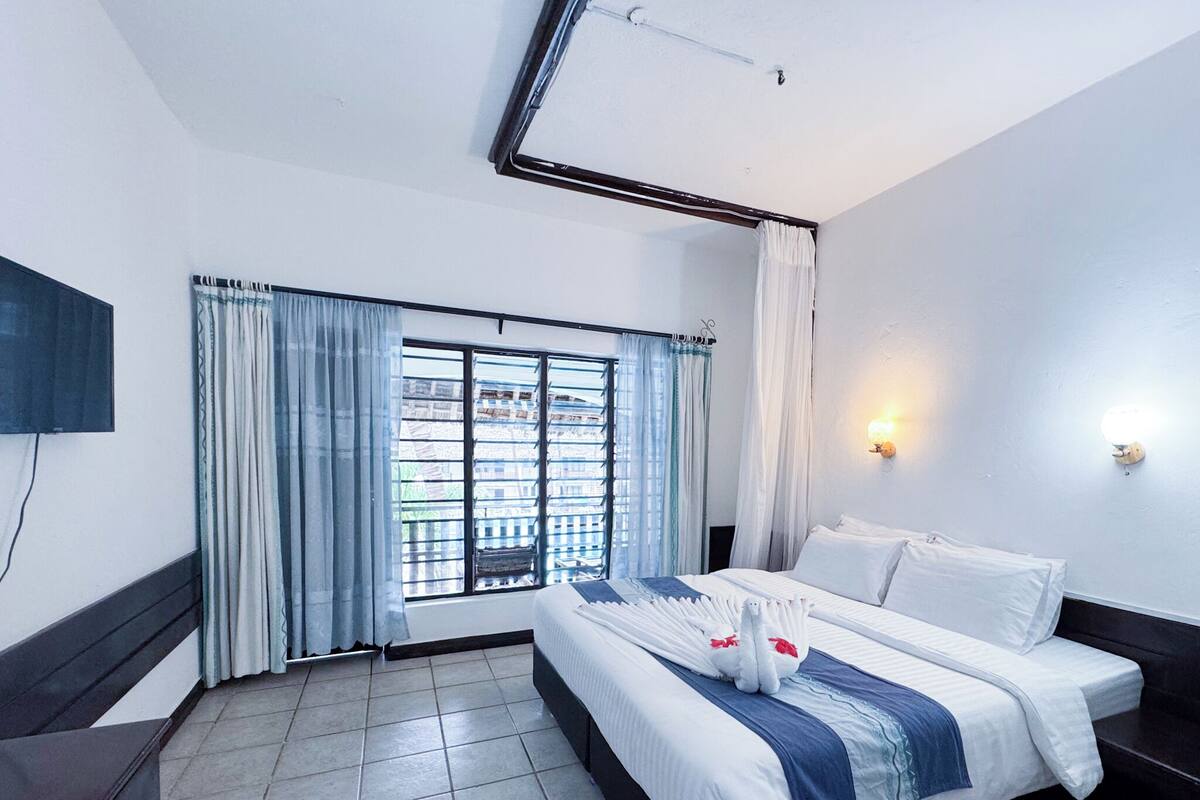 Quarto de hotel com cama, televisão, janela com cortinas e luminária fixada na parede.