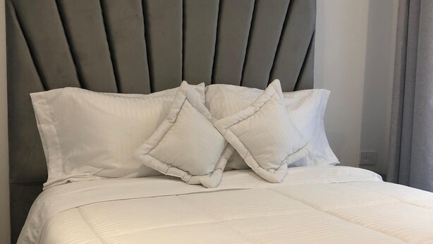 Cama bem arrumada com roupa de cama e almofadas brancas, cabeceira estofada cinza e parede lisa com uma janela à direita.
