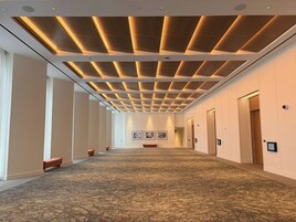 Corredor de hotel com carpete estampado, iluminação embutida e uma fileira de portas de um lado.