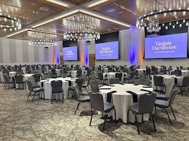 Uma sala de conferências com várias mesas e cadeiras, uma tela grande exibindo 'Virginia Guesthouse HOTEL & CONFERENCE CENTER', e um design moderno no teto com luminárias.
