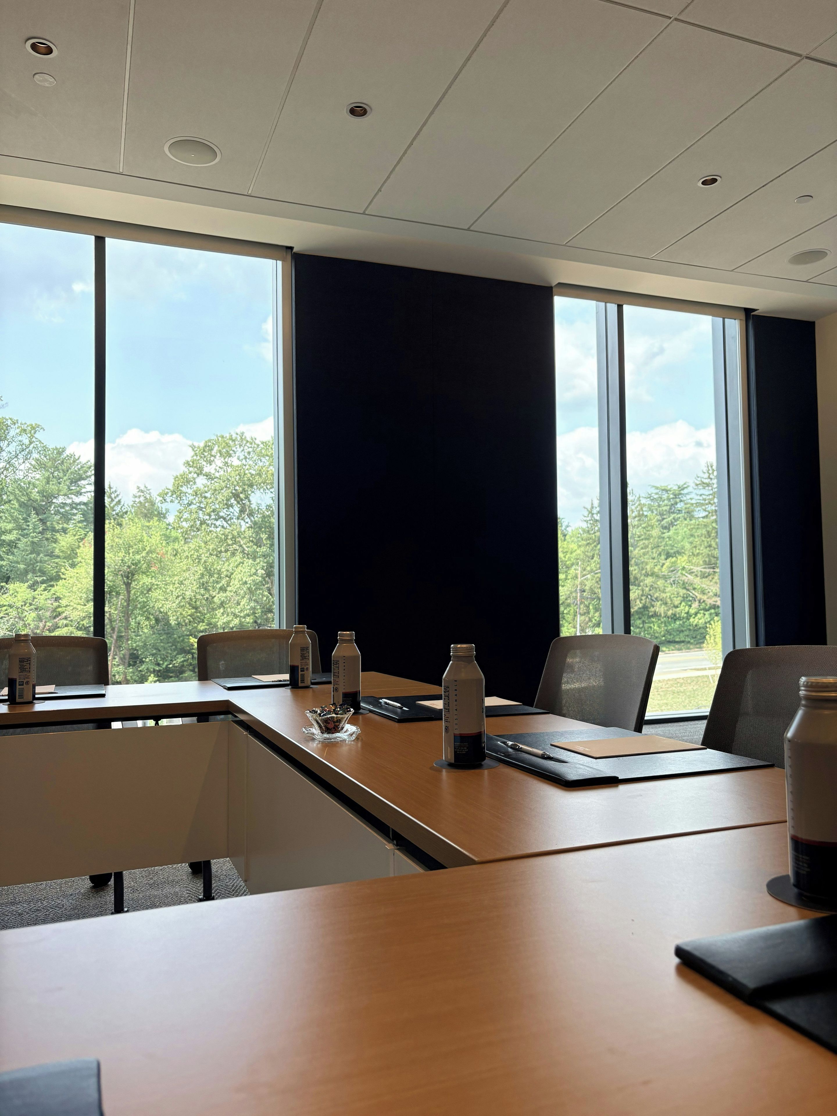 Sala de conferências com uma grande mesa, cadeiras e vista para árvores através de amplas janelas.