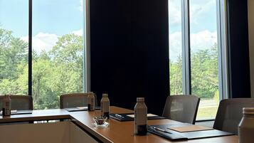 Sala de conferências com uma grande mesa, cadeiras e vista para árvores através de amplas janelas.