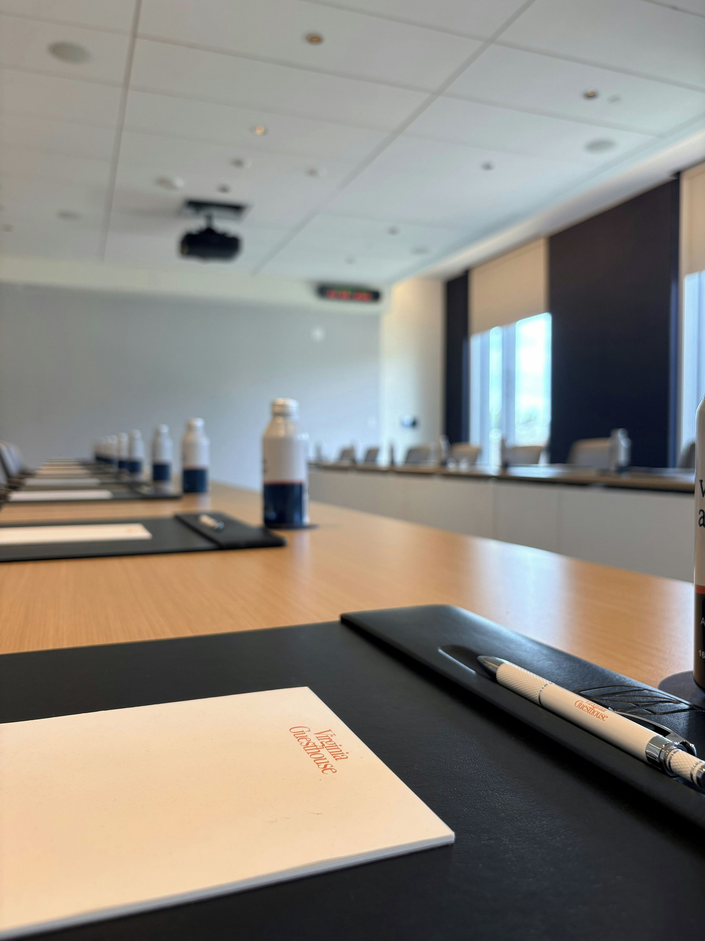 Sala de conferência com uma mesa comprida, garrafas de água, um laptop e uma caneta sobre um tapete preto.