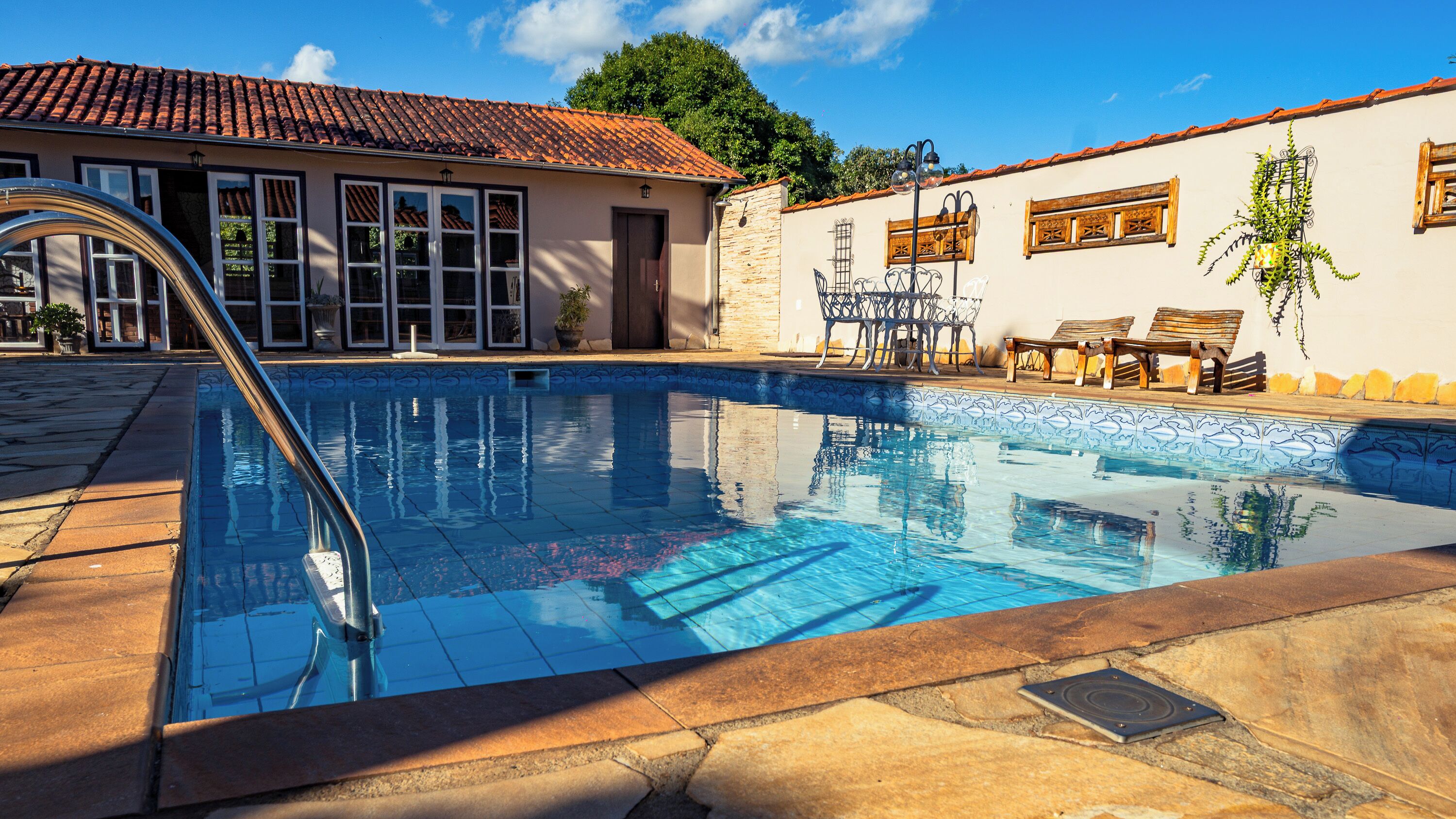 Uma piscina com deck ladrilhado, área de estar com cadeiras e mesa, e um edifício com janelas amplas e telhado ladrilhado.