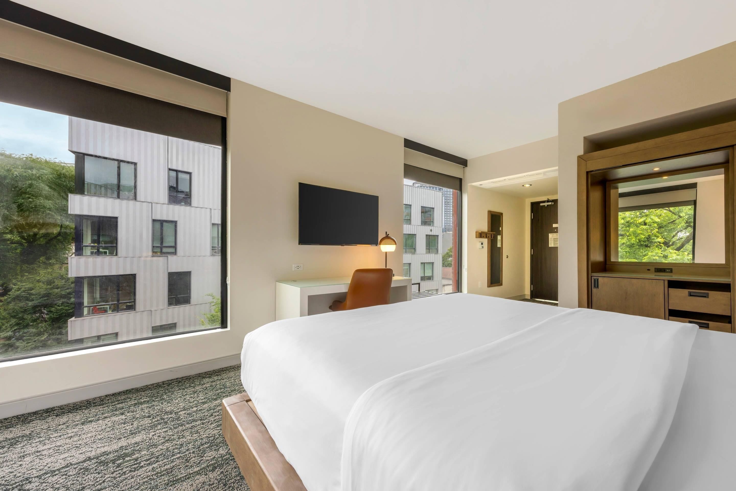 Quarto de hotel com uma cama grande, uma escrivaninha, uma cadeira, uma TV e uma janela com vista para edifícios e árvores.