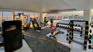 Academia em prédio alto com diversos equipamentos de ginástica, uma grande janela com vista para a cidade e um espaço amplo.