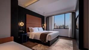 Quarto de hotel moderno com uma cama grande, mesa de cabeceira, escrivaninha e vista para a cidade.